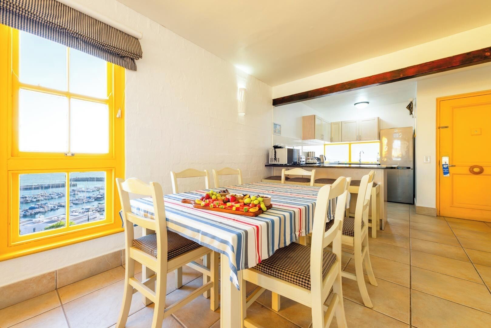 Uma área de refeições com uma mesa posta para quatro pessoas, uma cozinha ao fundo e vista para uma marina através de uma janela.