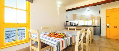 Uma área de refeições com uma mesa posta para quatro pessoas, uma cozinha ao fundo e vista para uma marina através de uma janela.