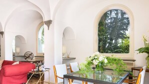 Uma área de refeições com mesa de vidro, cadeiras brancas, sofá vermelho e vista para árvores através de uma grande janela.