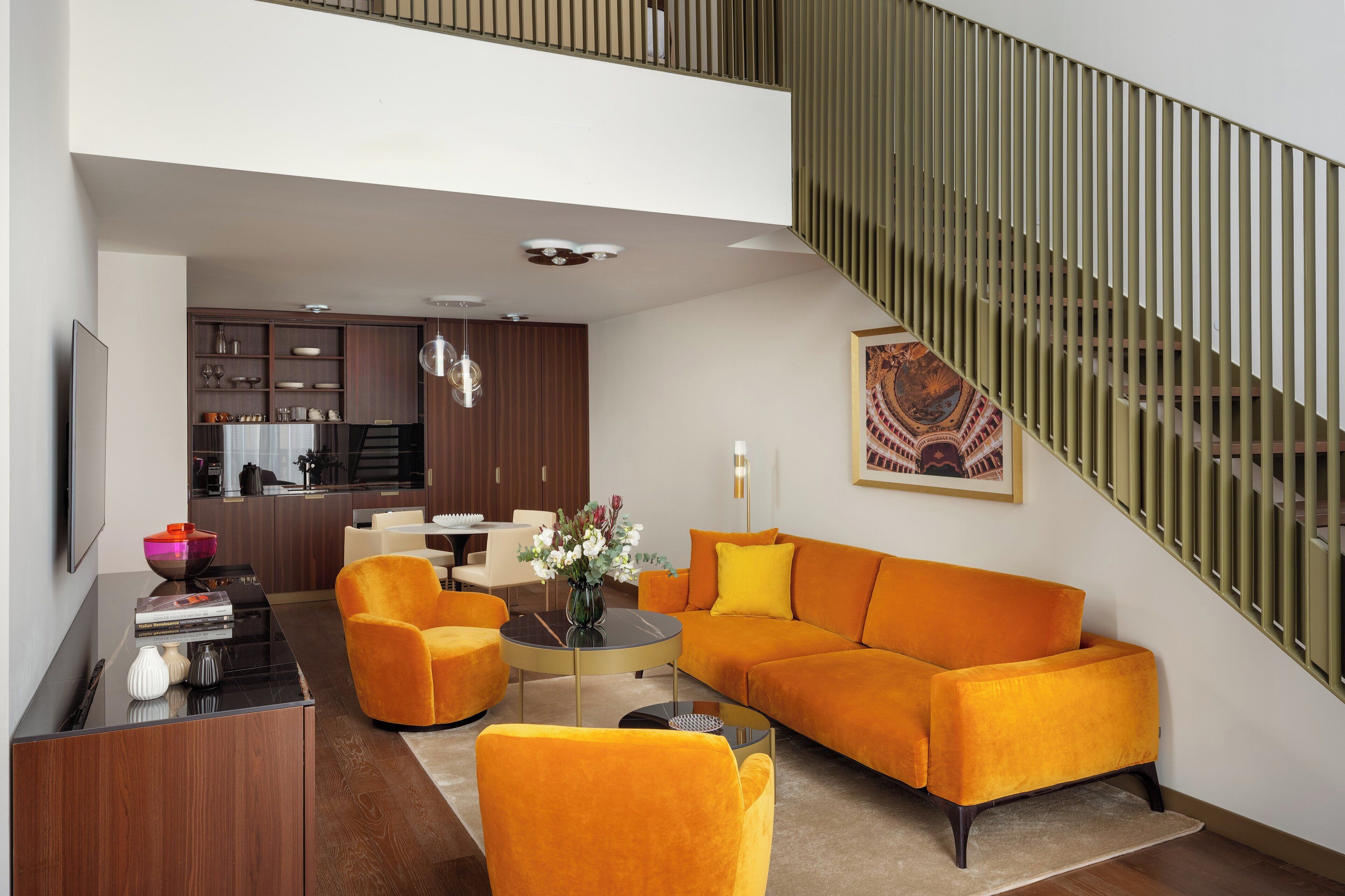 Uma sala de estar moderna com sofá, poltronas e mesa de centro. Uma cozinha aparece ao fundo e uma escada com corrimão.