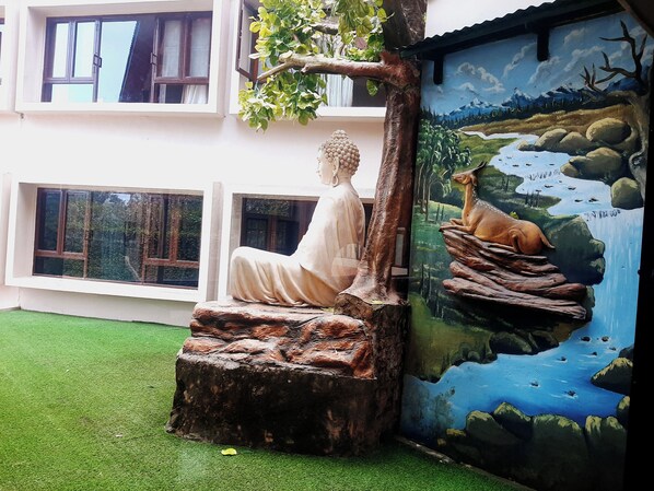 Estátua de uma figura sentada sobre um pedestal de rocha, uma árvore e um mural de paisagem com um veado.