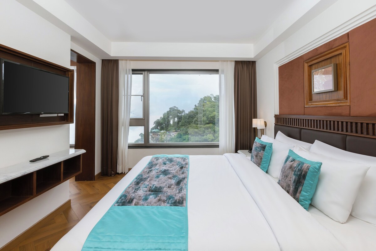 Quarto de hotel com uma cama grande, televisão de tela plana na parede, janela com vista para árvores e cabeceira de madeira.