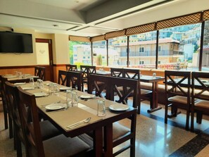 Uma sala de jantar com mesas preparadas para quatro pessoas, cadeiras de madeira e uma grande janela com vista para edifícios.
