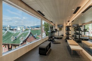 Uma academia com equipamentos de ginástica, bancos e uma grande janela com vista para um edifício tradicional.
