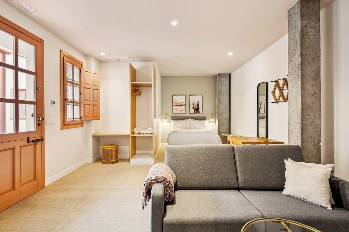 Quarto de hotel moderno com sofá cinzento, cama com roupa de cama branca, guarda-roupa de madeira e uma pequena mesa com abajur.