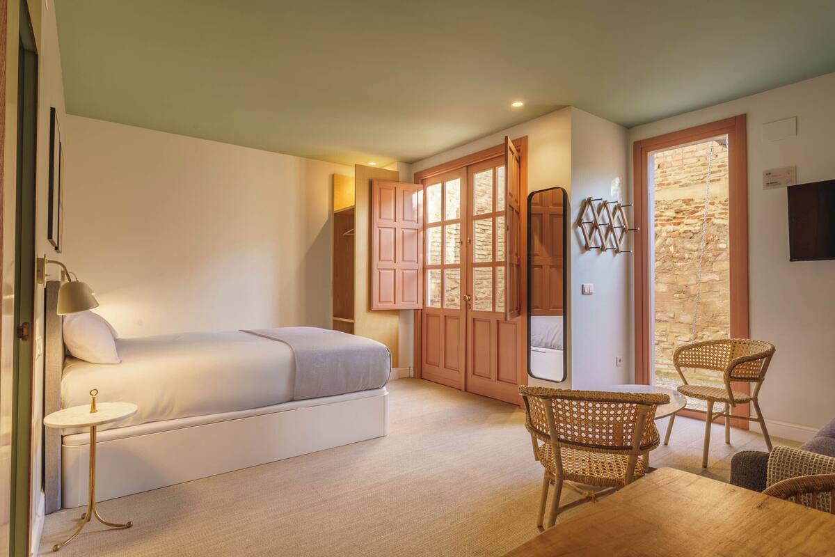 Um quarto com cama, uma mesinha, cadeiras de vime, espelho e uma parede de pedra.