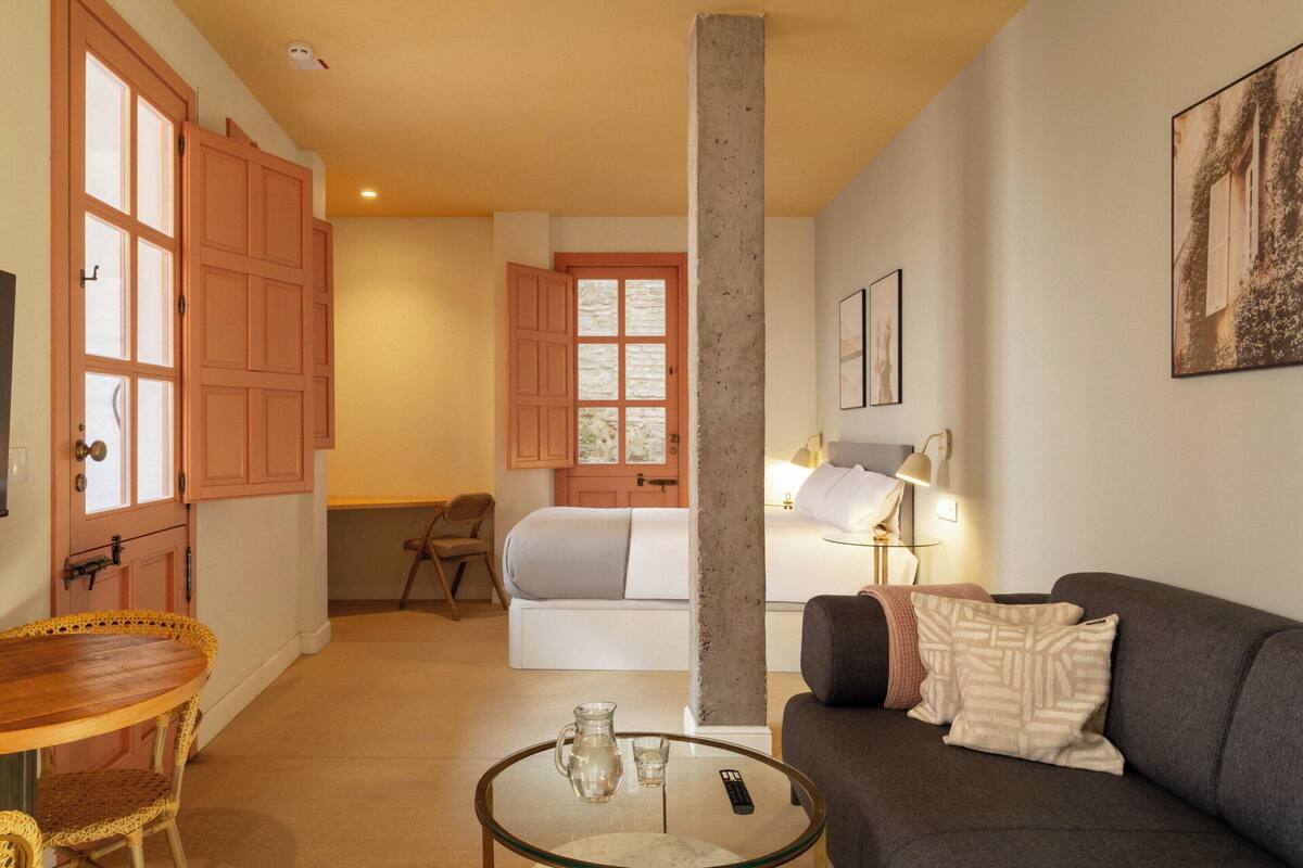 Quarto de hotel aconchegante com uma cama, um sofá, uma mesa de vidro com jarra e controle remoto, e uma cadeira.
