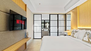 Quarto de hotel moderno com uma cama grande, uma TV presa a um painel de madeira, uma mesa de trabalho com luminária e uma porta de vidro deslizante que dá para uma varanda.