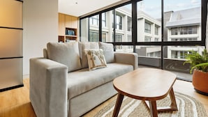 Sala moderna com sofá, mesa de centro de madeira e uma grande janela com vista para prédios.
