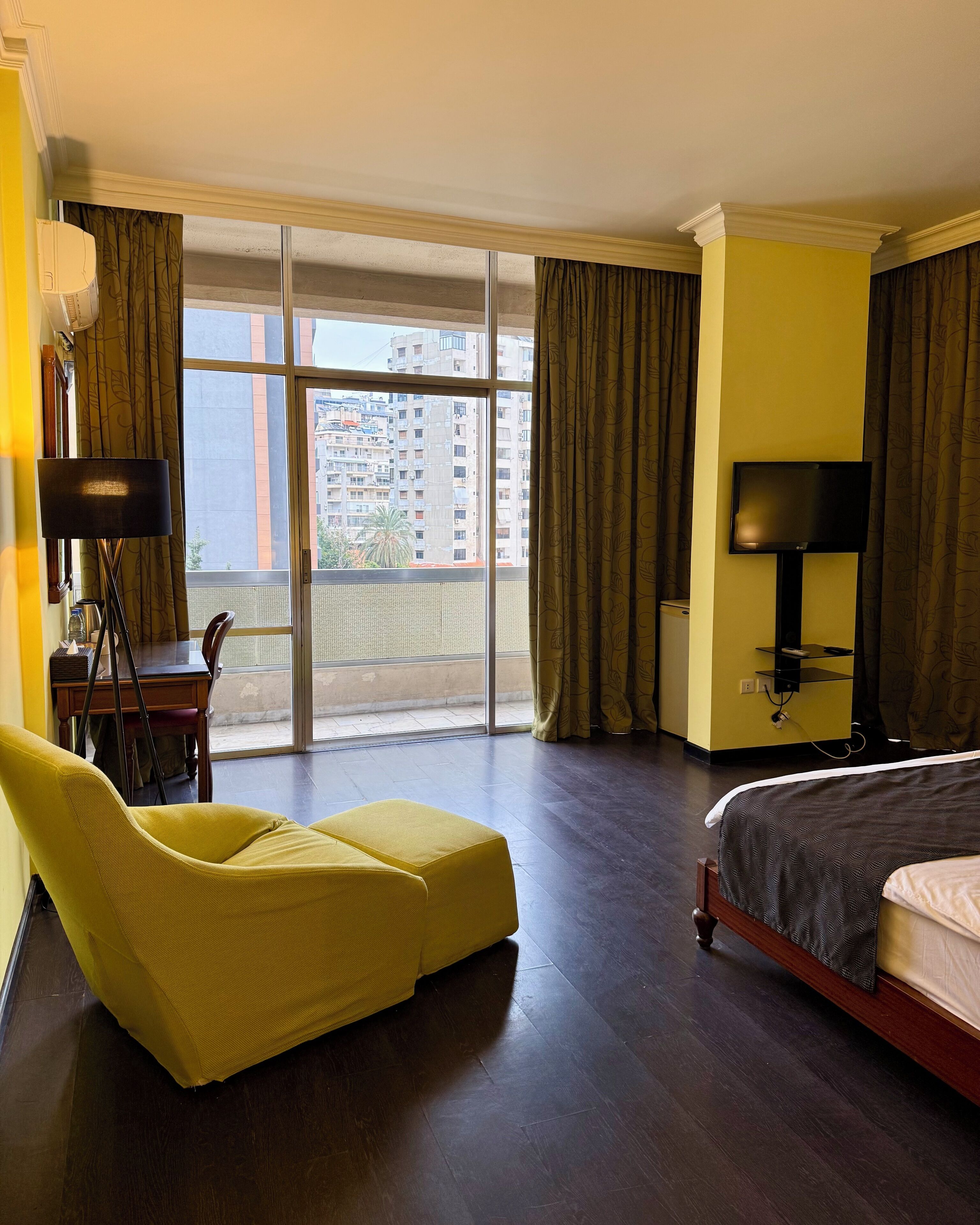 Quarto de hotel com uma cama, poltrona amarela, escrivaninha com luminária e televisão.