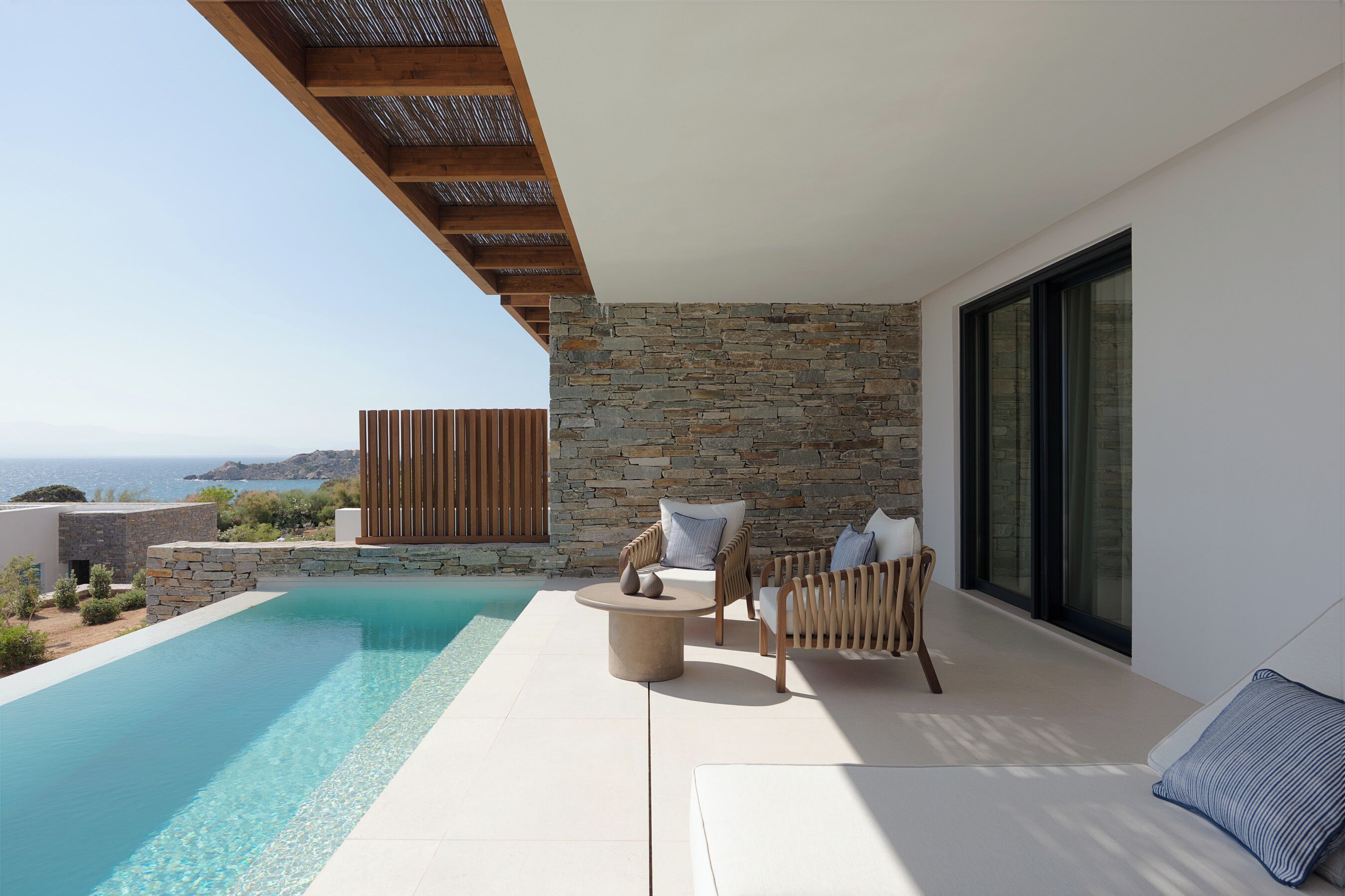 Uma área de estar moderna ao ar livre, com piscina, parede de pedra, móveis de madeira e vista para o mar.