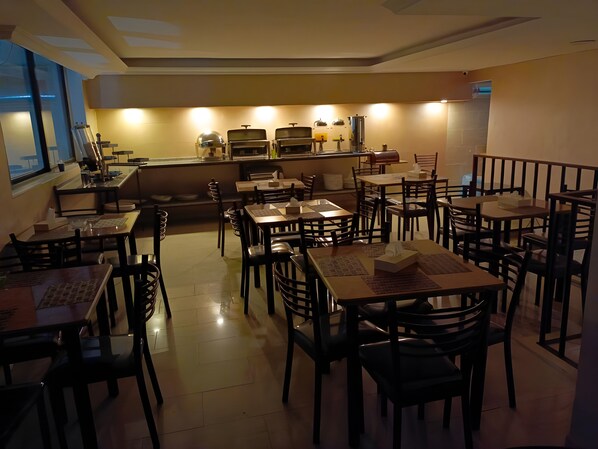 Um restaurante com várias mesas e cadeiras, um balcão com registadora de caixa e uma janela no lado esquerdo.