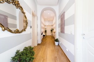Um corredor com piso de madeira, paredes brancas e um espelho grande e ornamentado à esquerda. Há plantas em vasos ao longo dos lados e uma porta no final.