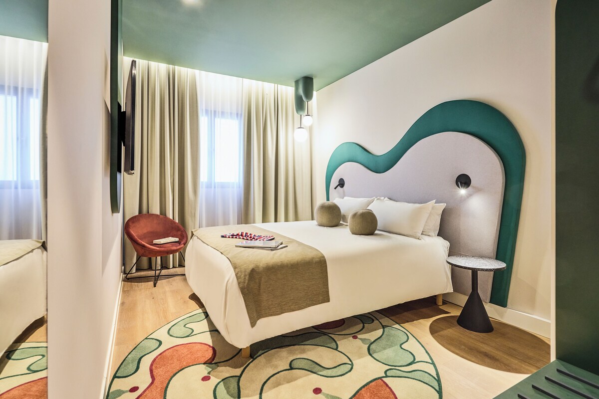 Quarto de hotel moderno com cama, uma cadeira, uma mesinha e uma janela grande com cortinas.