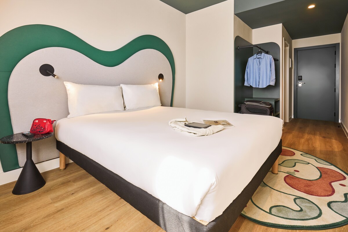 Uma cama bem arrumada com lençóis brancos, uma mesa de cabeceira preta com uma bolsa vermelha e uma cabeceira verde e branca.