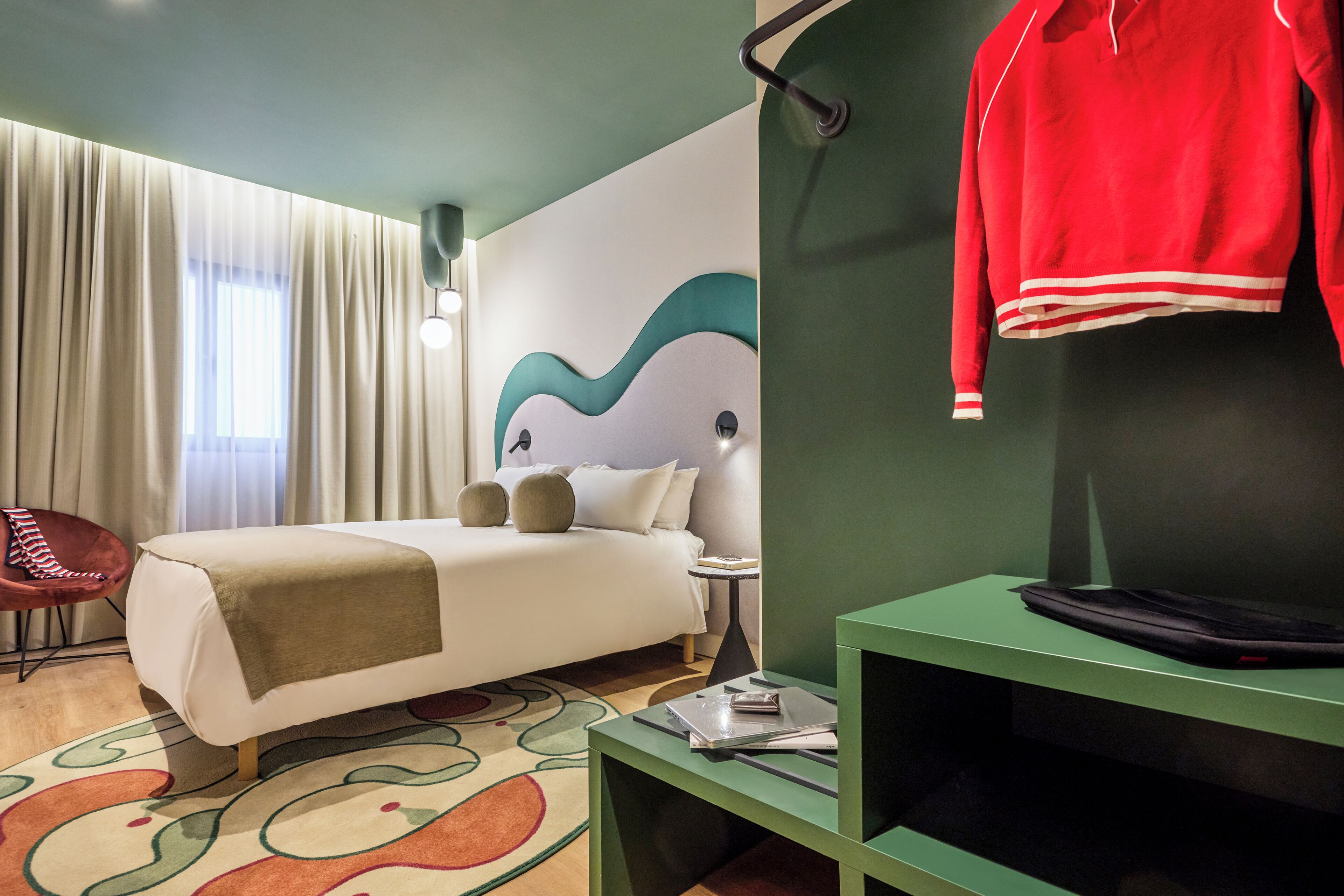 Quarto de hotel com cama, mesa de apoio verde, casaco vermelho pendurado na parede e janela com cortinas.