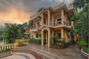 Um hotel com vários andares, varandas, um jardim com plantas e um caminho, e um céu limpo.