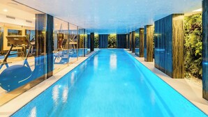 Piscina interior moderna com paredes de vidro, equipamentos de ginástica e vista para a paisagem urbana.