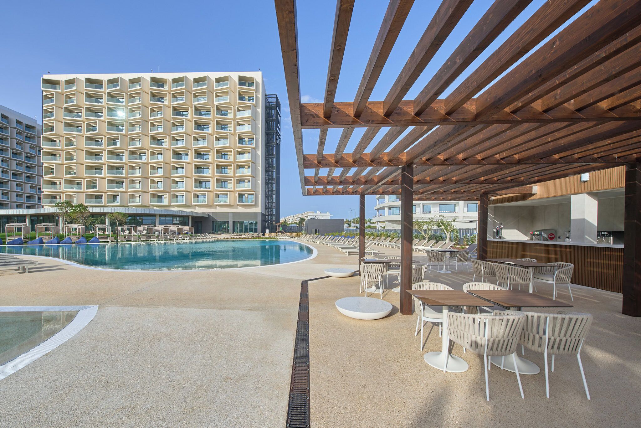 Uma área à beira da piscina com pergolado de madeira, espreguiçadeiras e piscina.