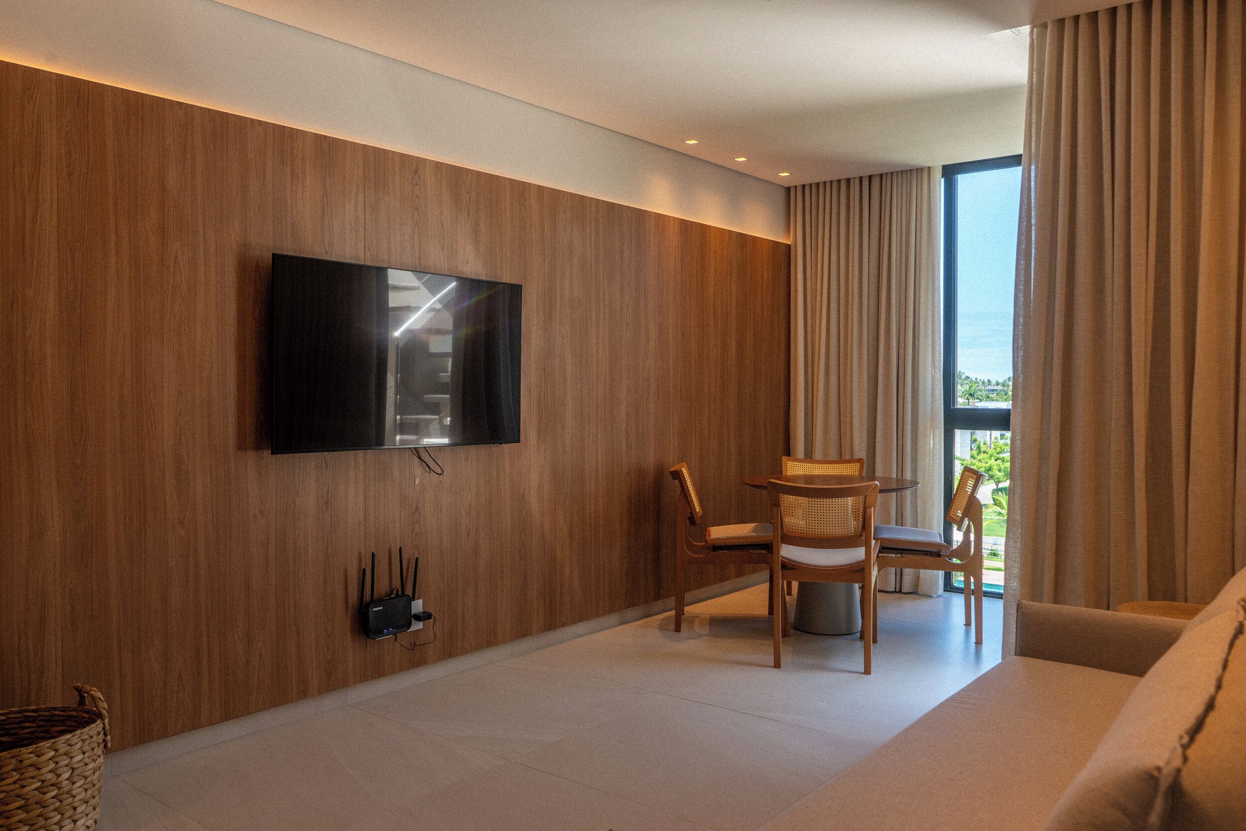 Uma sala de estar moderna com uma televisão de tela plana na parede, uma mesa redonda com cadeiras e uma grande janela com cortinas.