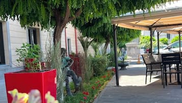 Uma área de estar coberta ao ar livre, com mesas e cadeiras, rodeada por vegetação e um vaso vermelho.