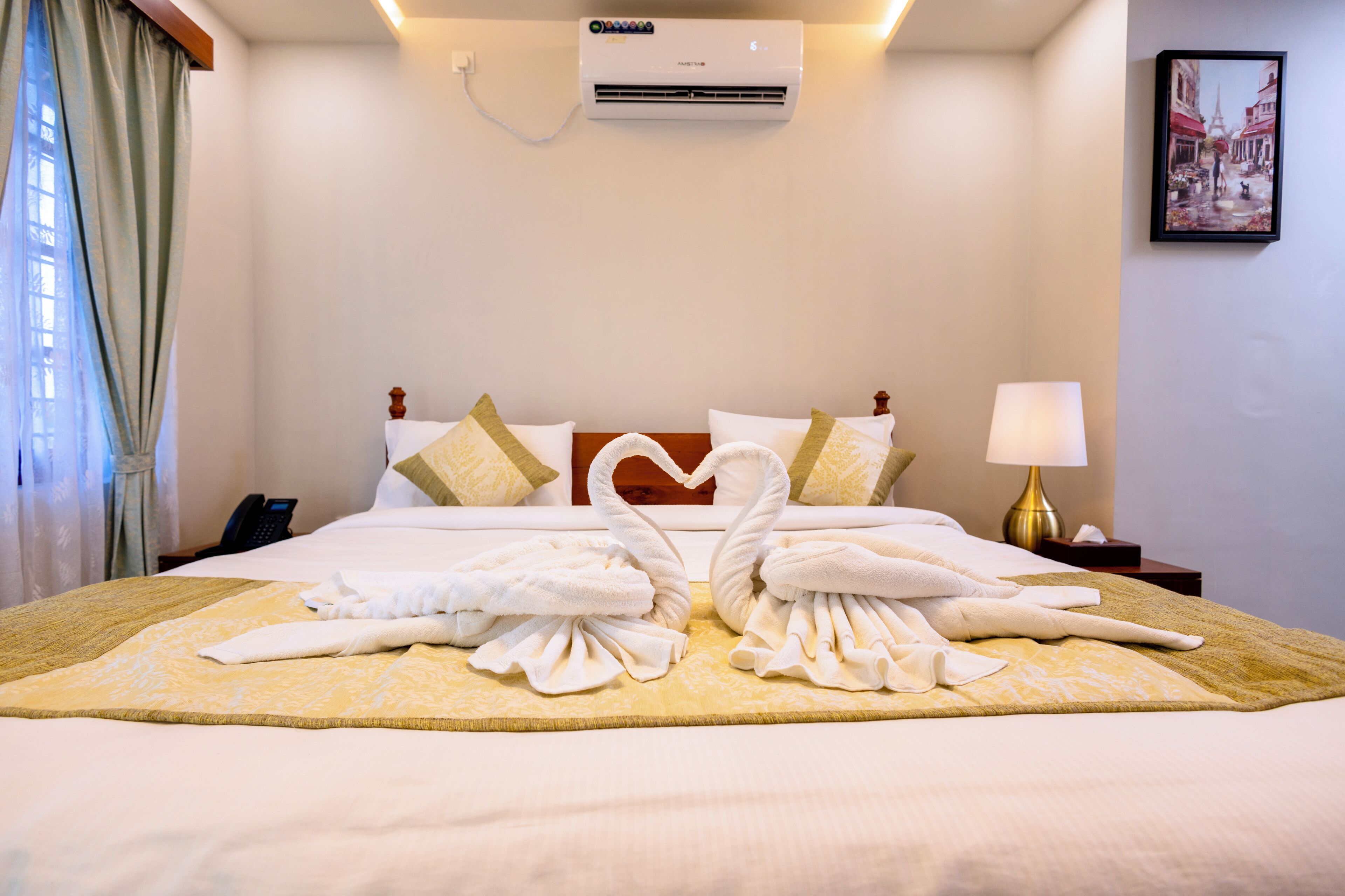 Quarto de hotel com uma cama bem arrumada, arranjo de toalhas em formato de coração, abajur e uma moldura na parede.