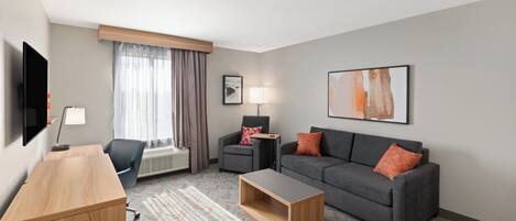 Quarto de hotel moderno com escrivaninha de madeira, sofá cinza e televisão de tela plana.