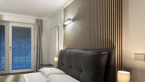 Quarto de hotel com uma cama grande, uma mesa de cabeceira com abajur, um radiador e uma janela com persianas.