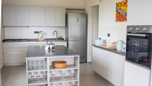 Cozinha moderna com armários brancos, geladeira de aço inoxidável, fogão a gás e forno embutido. Há taças de vinho e uma cesta sobre o balcão.