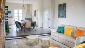 Uma sala de estar espaçosa com um sofá, uma mesa de jantar e cadeiras. É visível uma área de cozinha ao fundo.