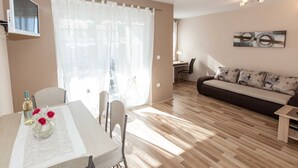 Uma sala de estar moderna com piso de madeira, um sofá, uma mesa de jantar com cadeiras, uma TV fixada na parede e uma janela grande com cortinas leves.
