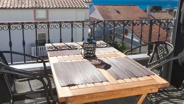 Uma mesa de madeira com dois jogos americanos trançados, um candeeiro e uma cadeira numa varanda com guarda de metal.