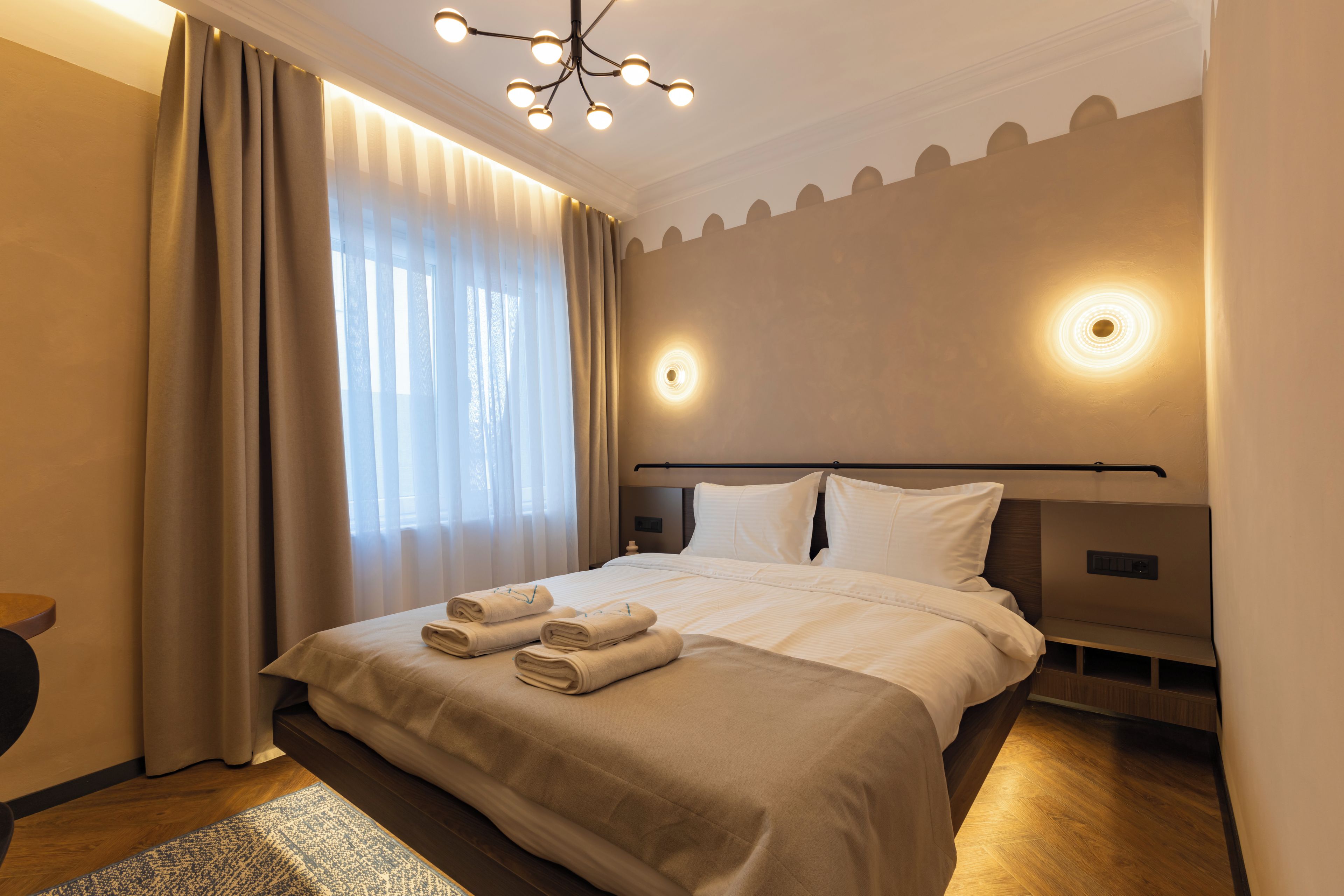 Quarto de hotel com cama grande, mesa de cabeceira, luminária na parede e janela com cortinas.