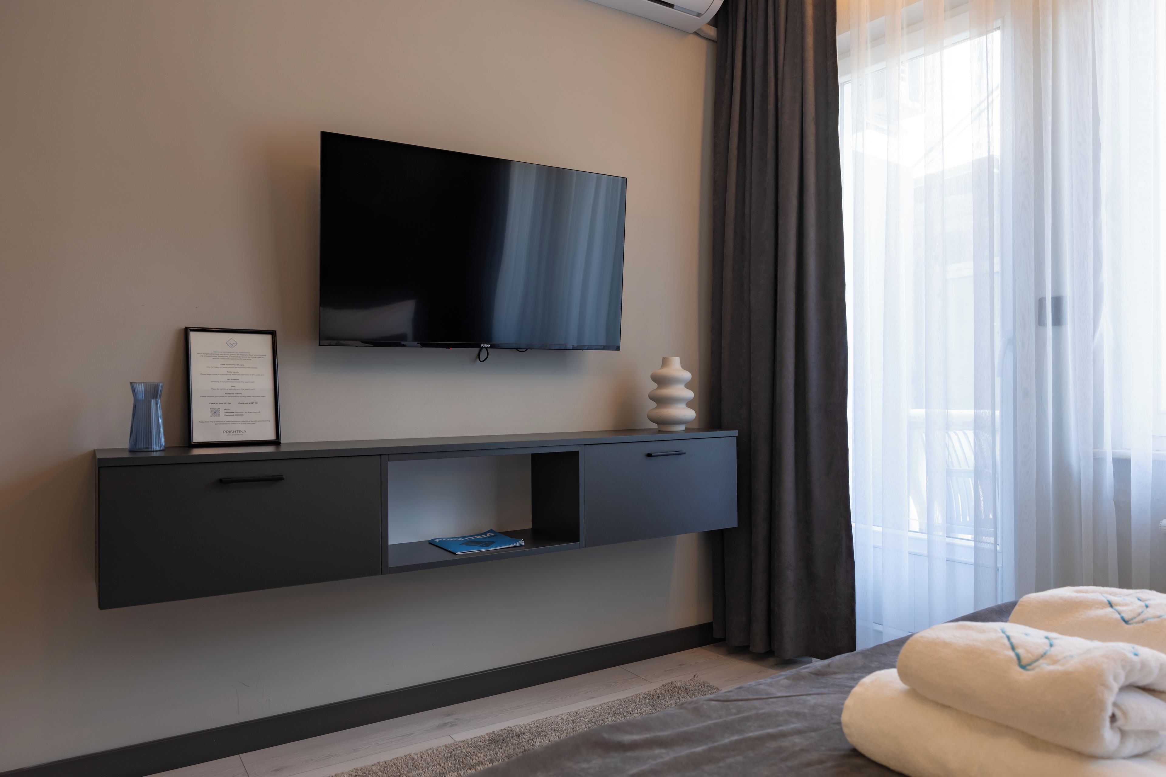 Quarto de hotel moderno com televisão de tela plana fixada em uma parede escura, mesa de cabeceira com um vaso e um certificado emoldurado, e uma janela grande com cortinas leves.