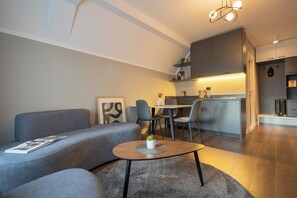 Uma sala de estar moderna com um sofá cinzento, uma mesa de centro e um espaço de refeições com cadeiras, e uma cozinha ao fundo.