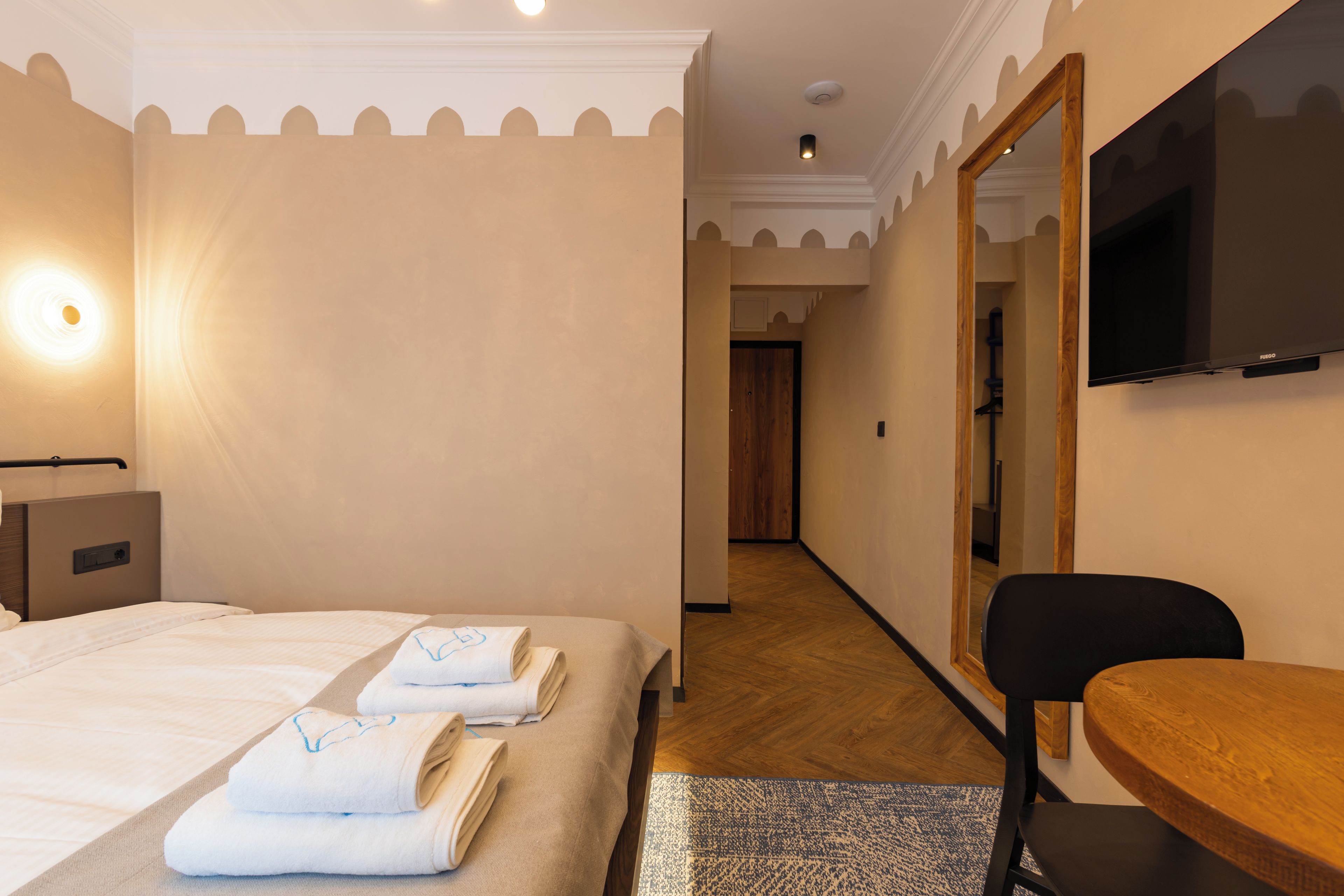 Quarto de hotel com cama, secretária, cadeira, televisão e banheiro ao fundo.