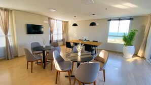 Uma zona de refeições moderna com uma mesa redonda, várias cadeiras, um vaso com planta e vista para o mar através de grandes janelas.