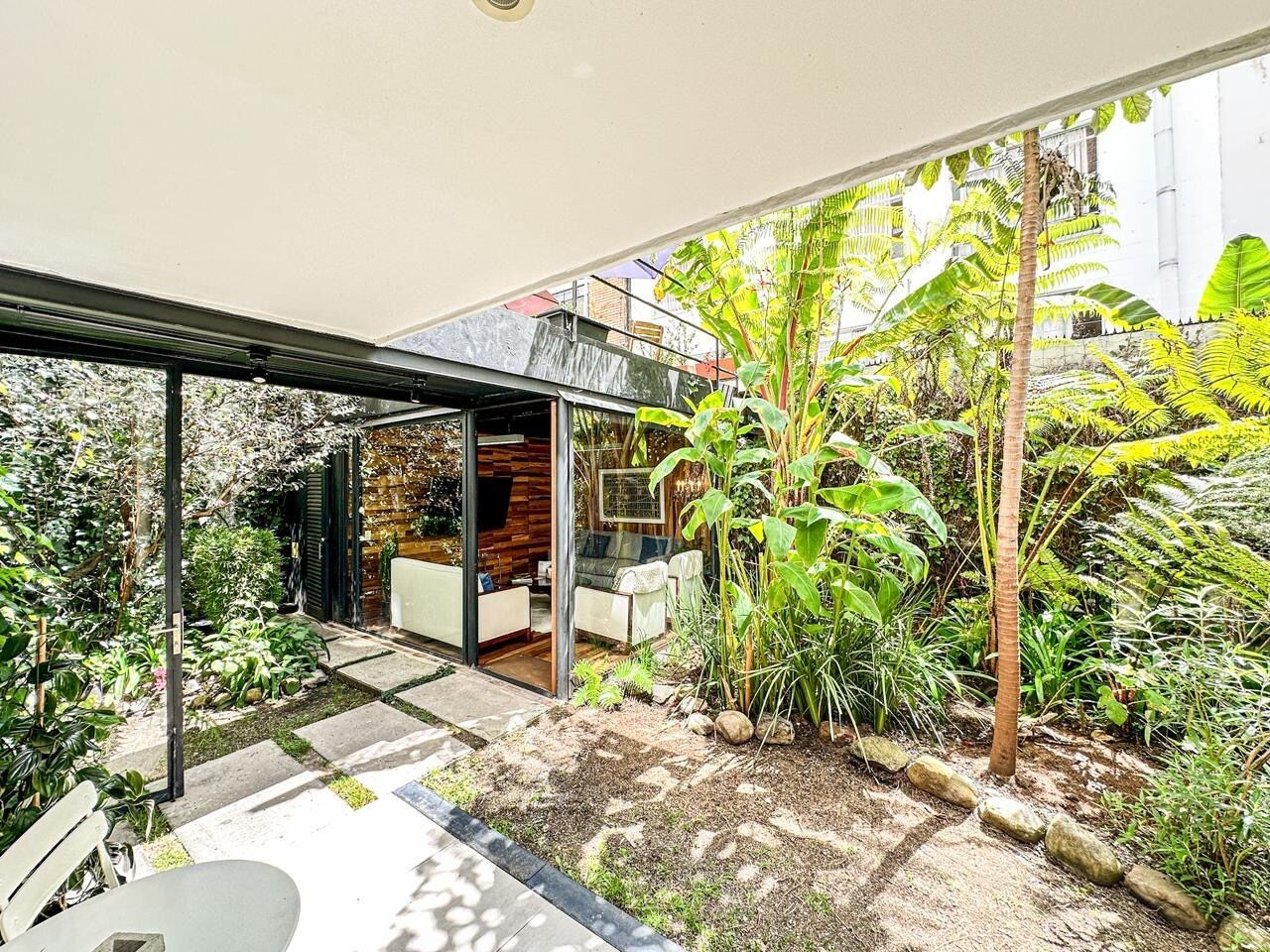 Uma casa moderna com porta de vidro que dá para uma sala de estar, cercada por vegetação exuberante e um caminho de pedra.