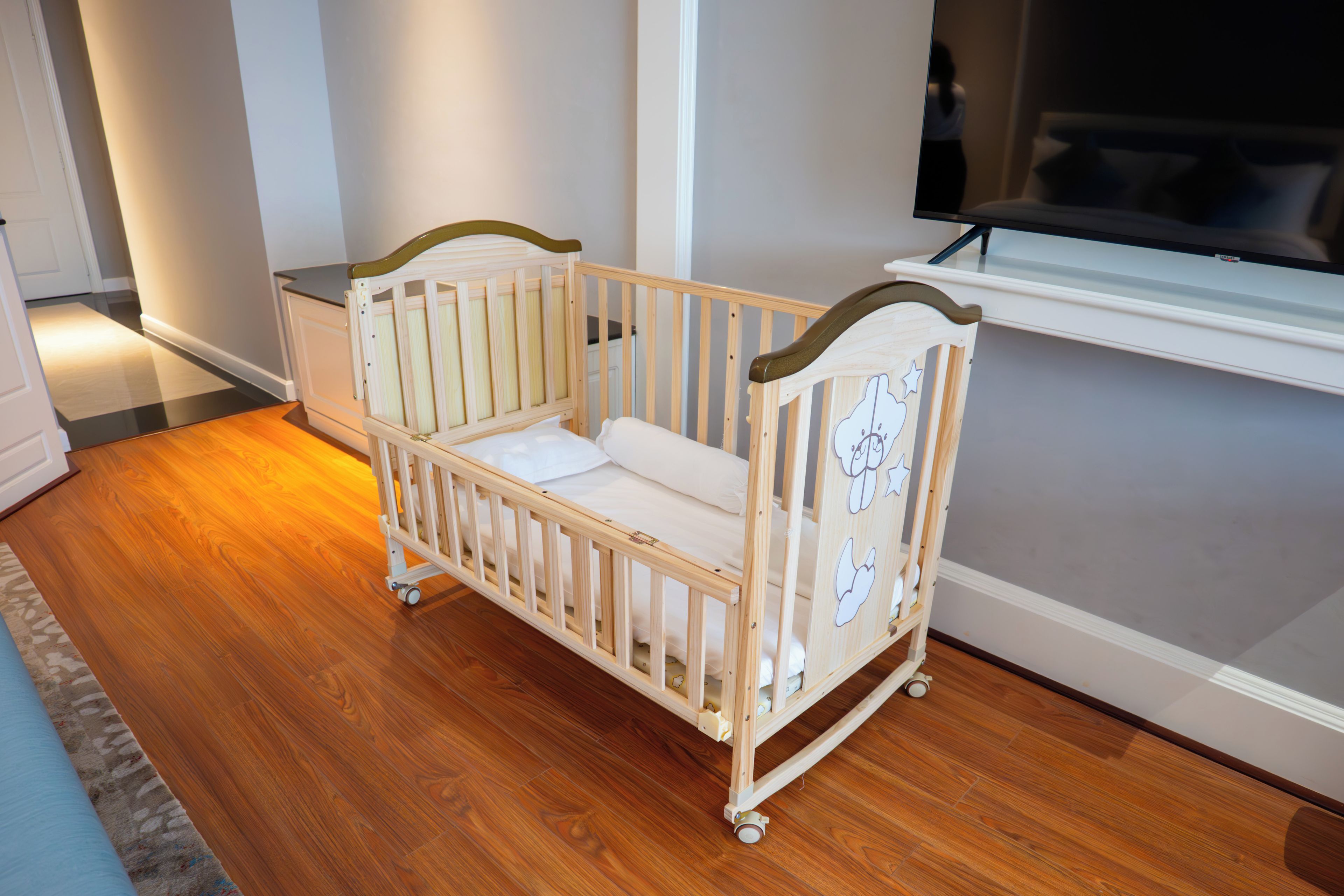 Berço branco com estrutura de madeira e cabeceira decorativa, colocado sobre um piso de madeira em um quarto com janela e parede branca.
