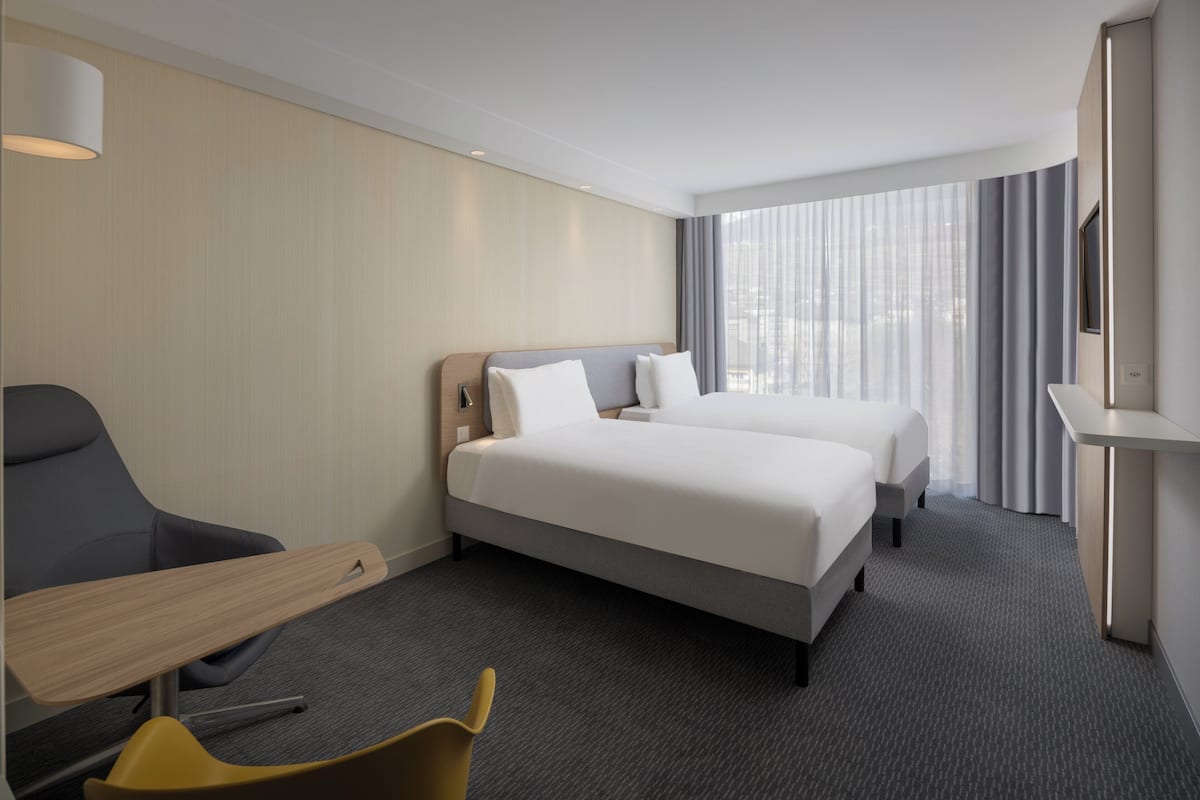 Quarto de hotel com duas camas, uma escrivaninha, uma cadeira e uma janela grande com cortinas.