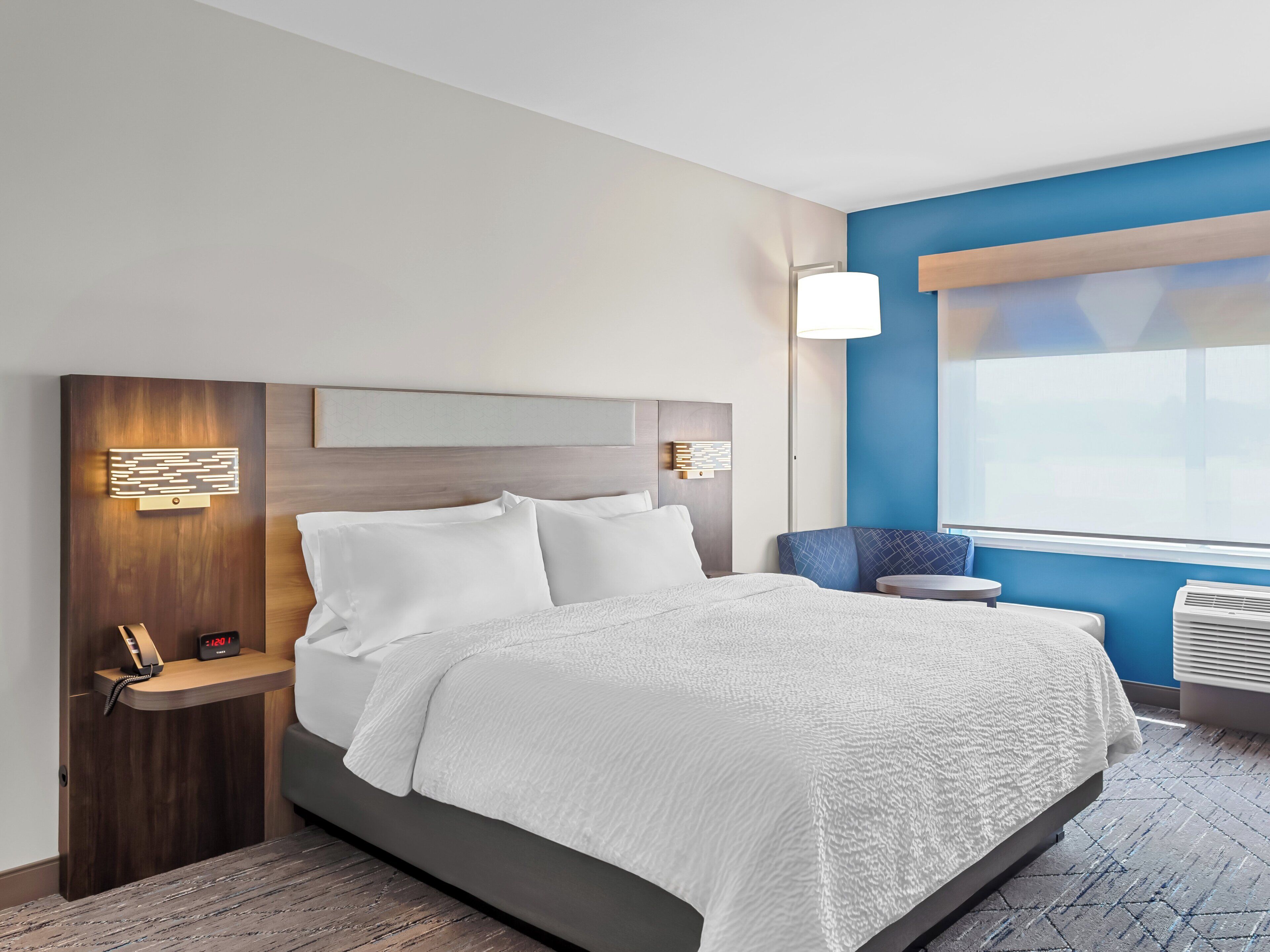 Quarto de hotel com uma cama grande, mesas de cabeceira, uma luminária de parede, uma pequena mesa redonda e uma janela com persianas.