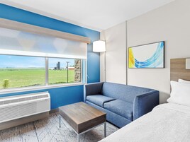 Quarto de hotel moderno com uma janela grande, um sofá azul, uma mesa de centro de madeira e uma cama com roupa de cama branca.