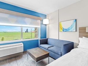 Quarto de hotel moderno com uma janela grande, um sofá azul, uma mesa de centro de madeira e uma cama com roupa de cama branca.