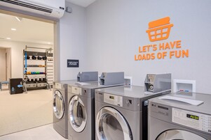 Lavanderia com máquinas de lavar e secar industriais, sinal indicando produtos de lavanderia disponíveis e um aviso na parede sobre diversão garantida.