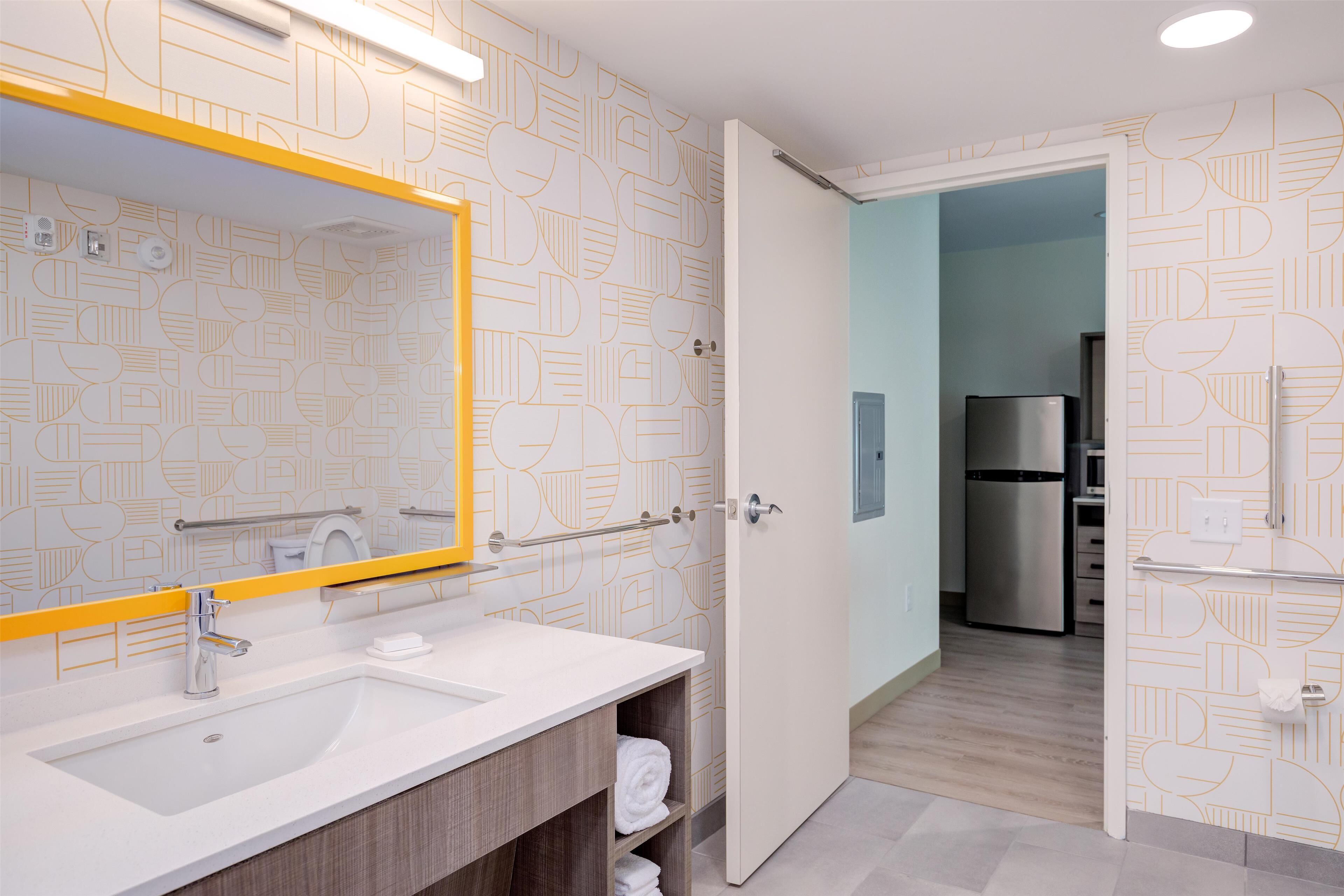 Uma casa de banho moderna com um espelho grande, lavatório branco e toucador em madeira. Uma porta dá acesso a uma zona de cozinha com frigorífico.