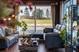 Uma sala de estar moderna com sofá, poltrona e mesa de centro, grandes janelas com vista para uma área gramada e árvores, e um armário de vidro com objetos decorativos.