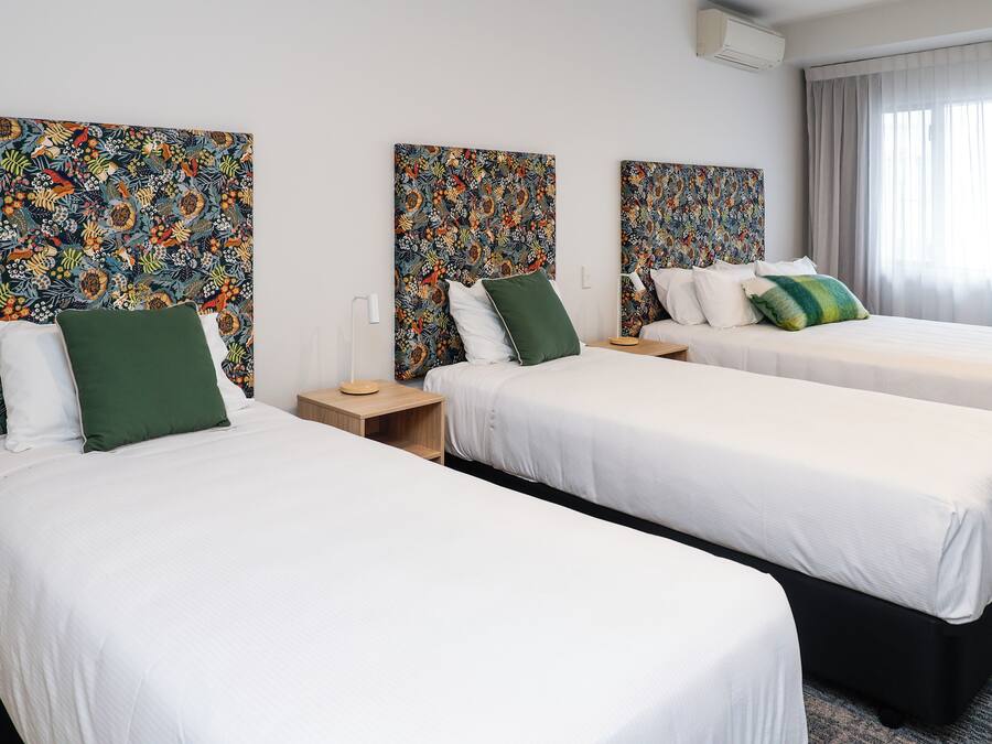 Quarto de hotel com três camas, cada uma com roupa de cama branca e almofadas verdes. Apresenta grandes cabeceiras com padrão floral e uma janela ampla com cortinas leves.