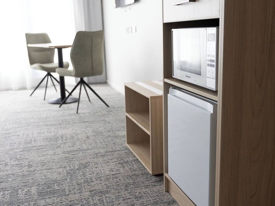 Quarto de hotel moderno com cozinha compacta, carpete cinza, escrivaninha de madeira e uma cadeira cinza.