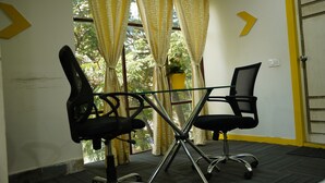Uma sala de reuniões com duas cadeiras de escritório pretas, uma mesa de vidro e uma janela com cortinas.