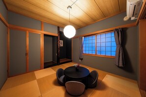 Um quarto japonês tradicional com tatames, uma mesa redonda com cadeiras, um ventilador de teto e uma janela com cortinas azuis.